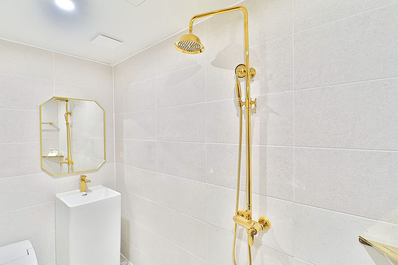 Casa de banho com sistema de chuveiro dourado, lavatório branco e espelho com moldura dourada.