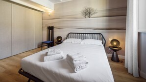 Cama bem arrumada com roupa de cama e toalhas brancas, mesa de cabeceira com abajur, e uma cadeira em quarto moderno com piso de madeira e prateleira na parede.
