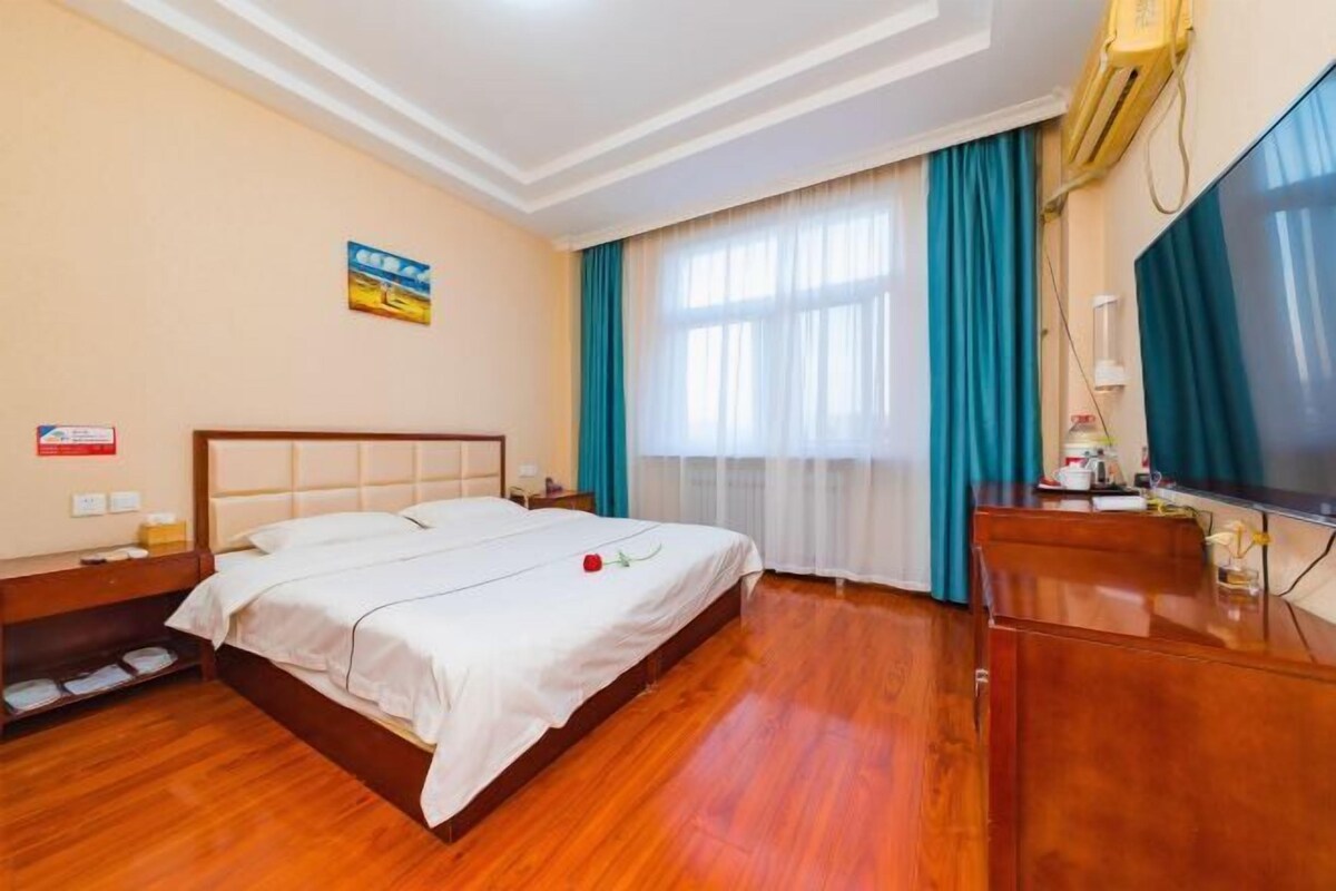 Quarto de hotel com piso de madeira, cama com roupa de cama branca, televisão em suporte de madeira e janela com cortinas.