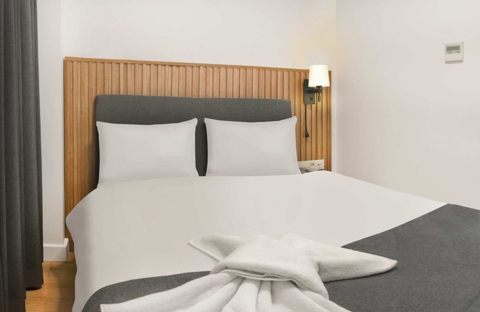 Quarto de hotel com cabeceira de madeira, roupa de cama branca, luminária de cabeceira e termostato na parede.