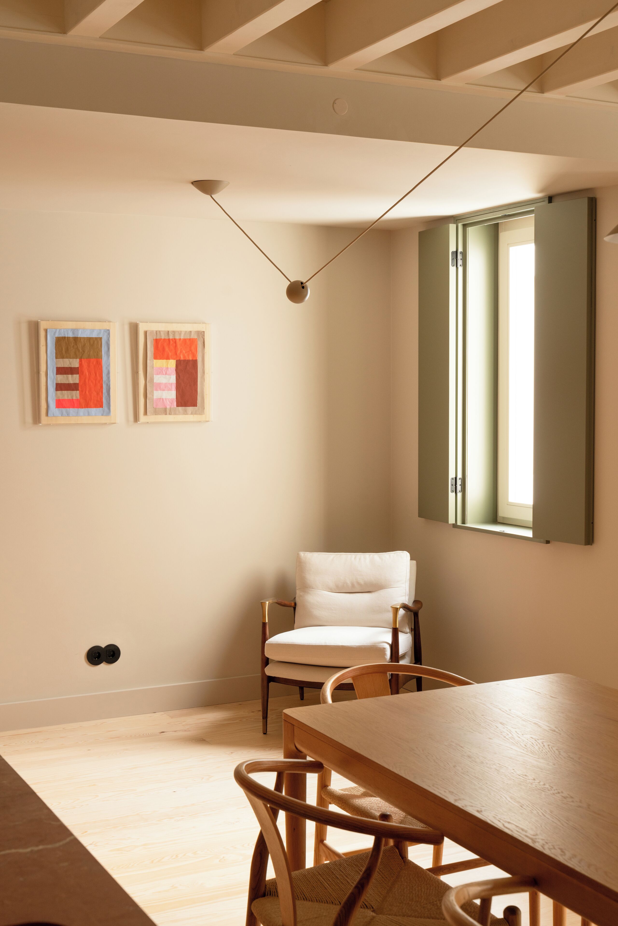 Um quarto com uma mesa de jantar de madeira, uma cadeira branca e uma janela com persianas verdes. Há duas obras de arte emolduradas na parede.