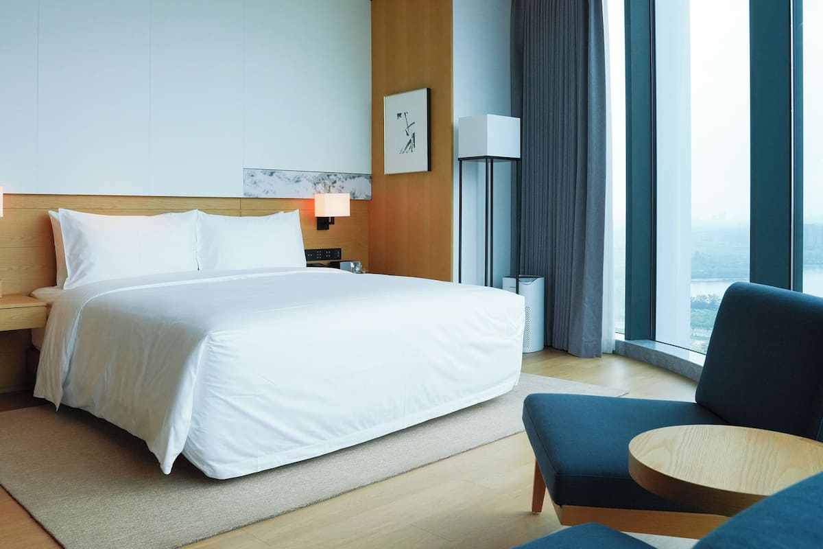 Quarto de hotel moderno com uma cama grande, mesas de cabeceira com luminárias, vista da cidade e uma poltrona azul.