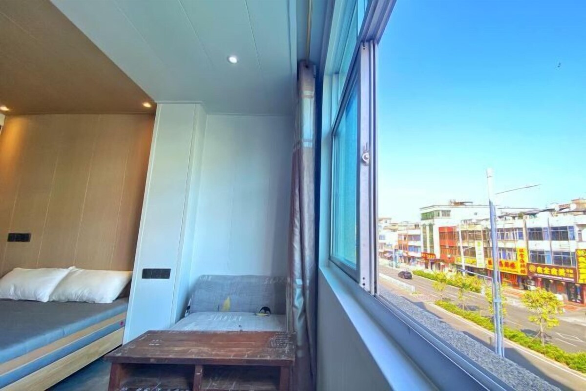 Quarto com cama, uma mesinha e janela com vista para a cidade.