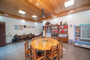 Quarto com teto de madeira, mesa redonda com cadeiras, geladeira e prateleiras com diversos objetos.