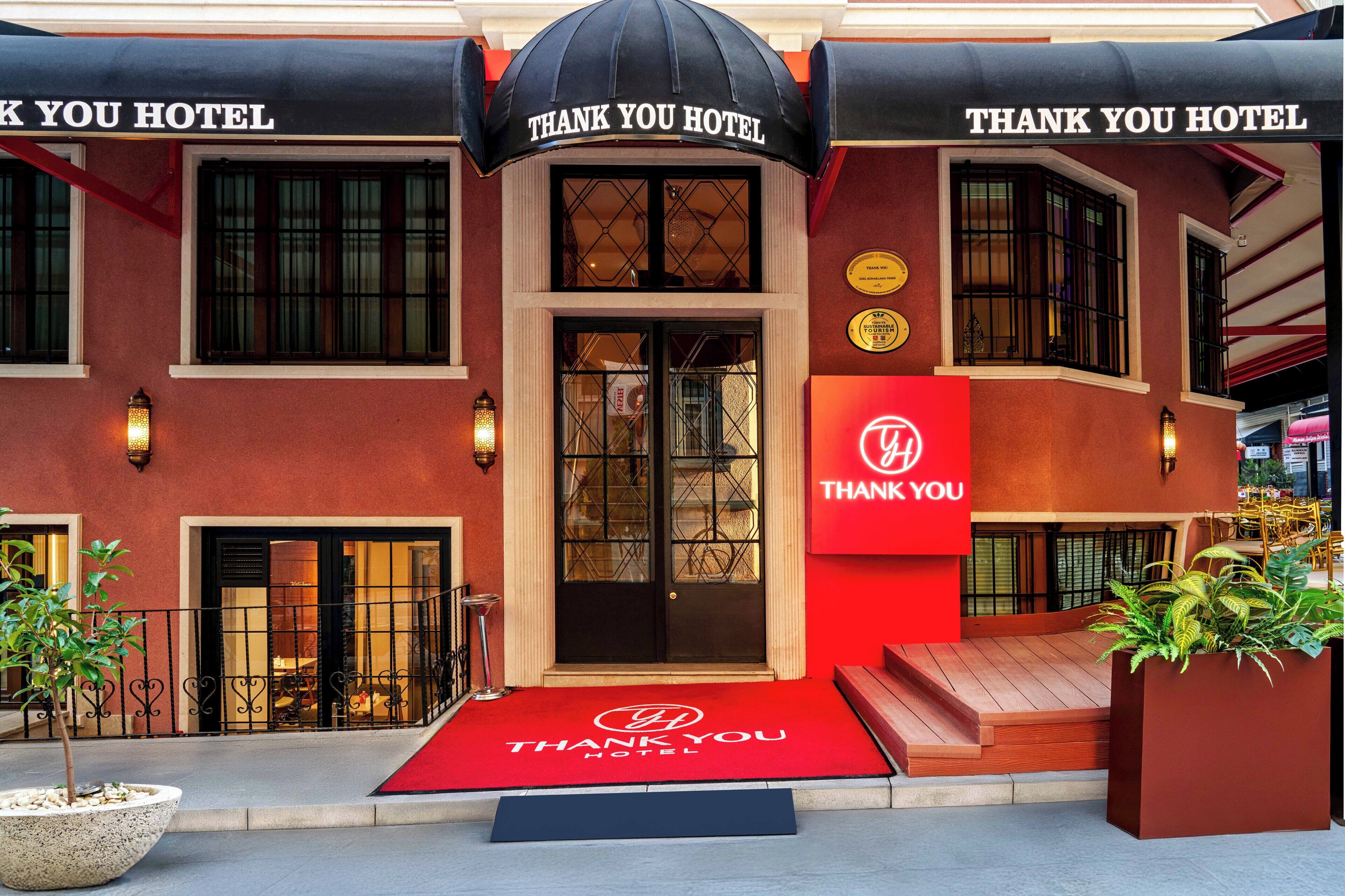 Entrada de hotel com tapete vermelho, toldo preto e placa com a inscrição 'THANK YOU HOTEL'.