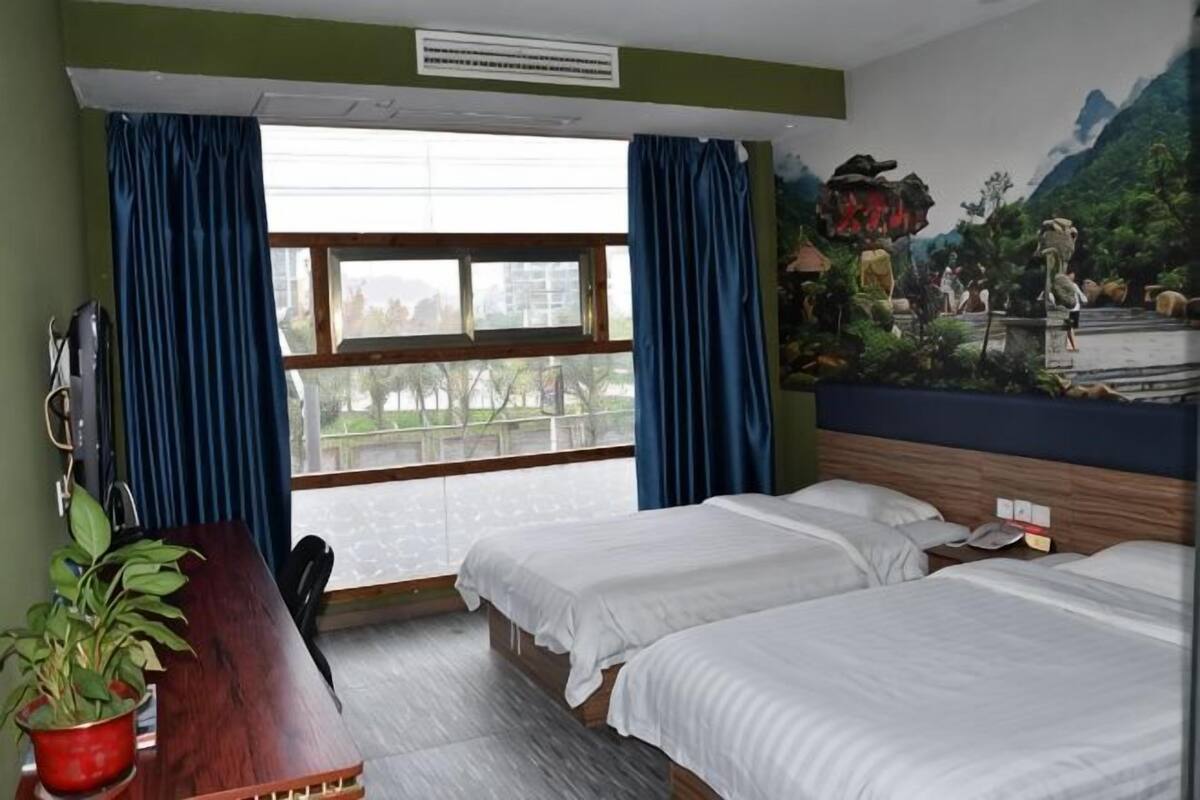 Quarto de hotel com duas camas, uma mesa com uma planta, uma janela com cortinas e um mural na parede.