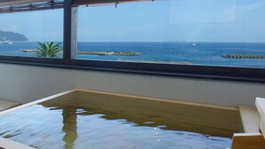 Quarto com grande janela com vista para um mar tranquilo, uma mesa pequena com superfície refletora e uma planta em vaso.