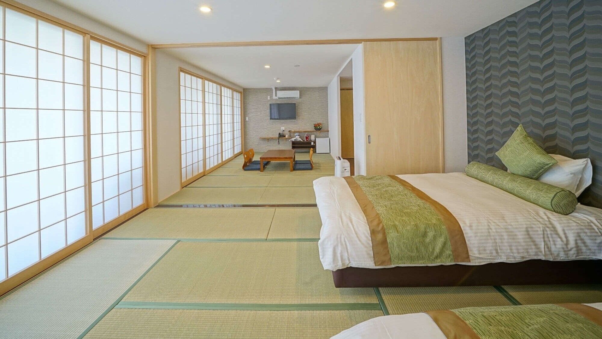 Quarto japonês tradicional com tatames, uma cama com detalhes verdes, uma escrivaninha pequena e uma televisão.
