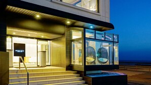 Uma entrada de hotel moderna com fachada de vidro, escadaria que leva à entrada e área de piscina visível do lado de fora.