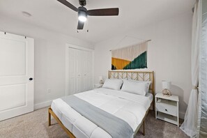 Quarto com cama, ventoinha no teto, porta branca, mesa de cabeceira branca e tapeçaria na parede.