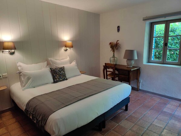 Quarto com cama, duas luminárias na parede, escrivaninha com cadeira e janela com vista para o verde.