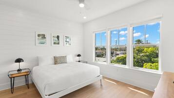 Quarto com cama, duas mesas de cabeceira, ventilador de teto e janelas amplas com vista para a cidade.