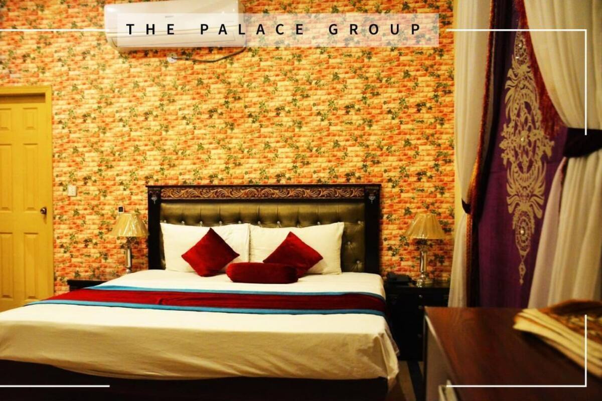 Quarto de hotel com parede em padrão de tijolos, porta de madeira, cama com roupa de cama branca e almofadas vermelhas, e mesa de cabeceira com abajur.