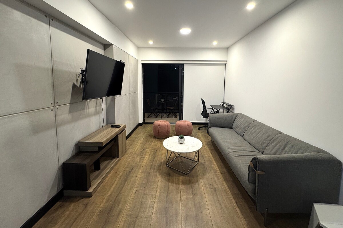 Uma sala de estar moderna com um sofá cinzento, uma mesa de centro redonda, uma TV de tela plana na parede e um banco de madeira.
