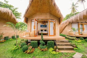 Uma cabana com telhado de palha e área de estar central, cercada por vegetação exuberante e um pequeno jardim.