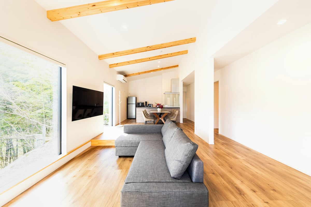 Uma sala de estar moderna com um sofá seccional cinza, vigas de madeira no teto e uma grande janela com vista para o verde.
