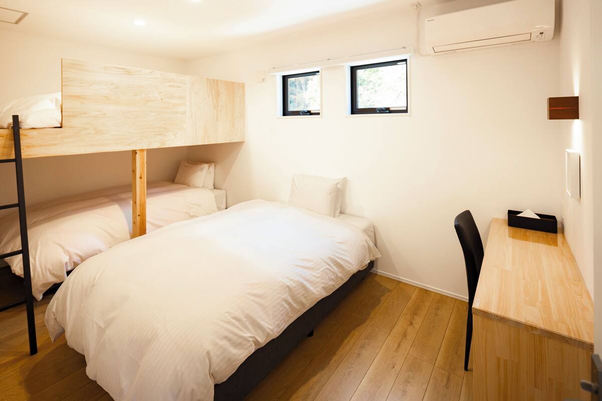 Quarto em dormitório com beliches, uma escrivaninha e duas janelas.