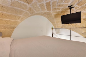 Quarto com teto de pedra, cama com colcha bege e televisão de tela plana montada na parede.