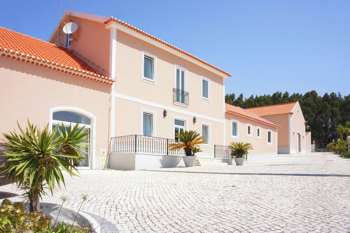 Um edifício de dois andares com telhado em tijolos, pátio pavimentado e plantas em vasos.