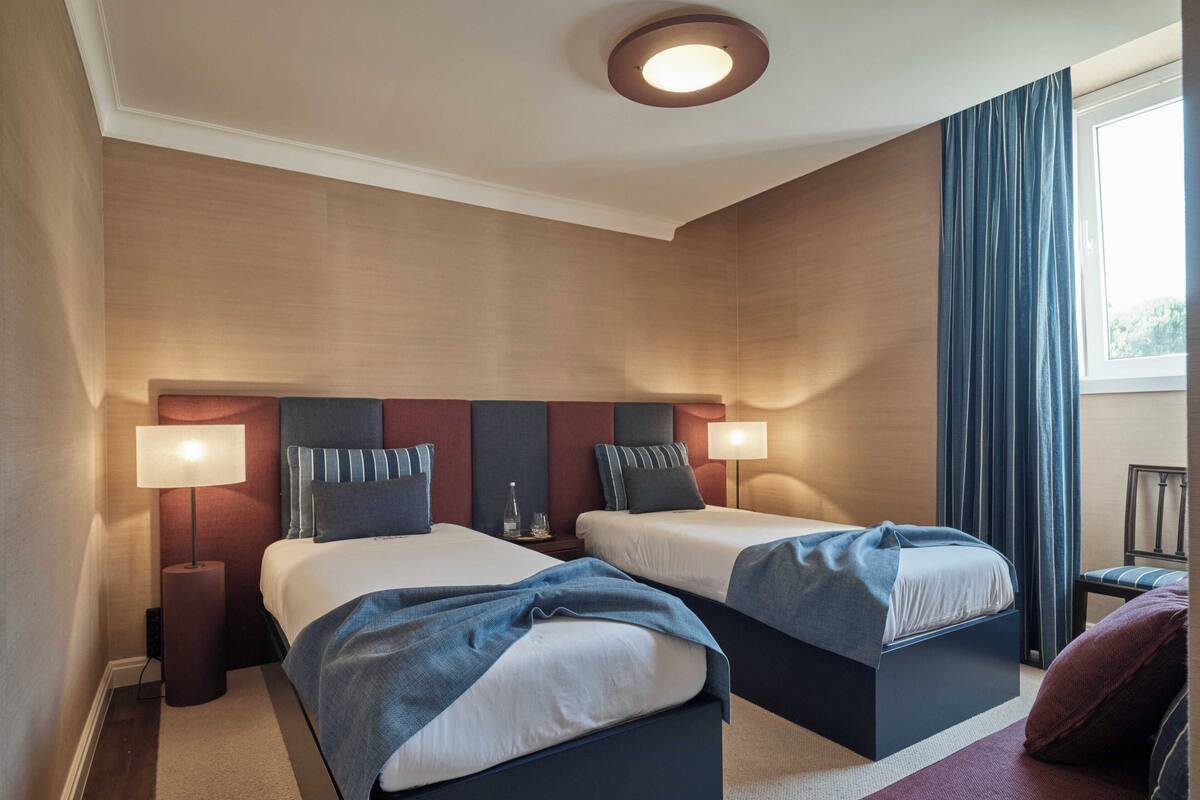 Quarto de hotel com duas camas, cada uma com cabeceira, abajures e uma janela com cortinas.
