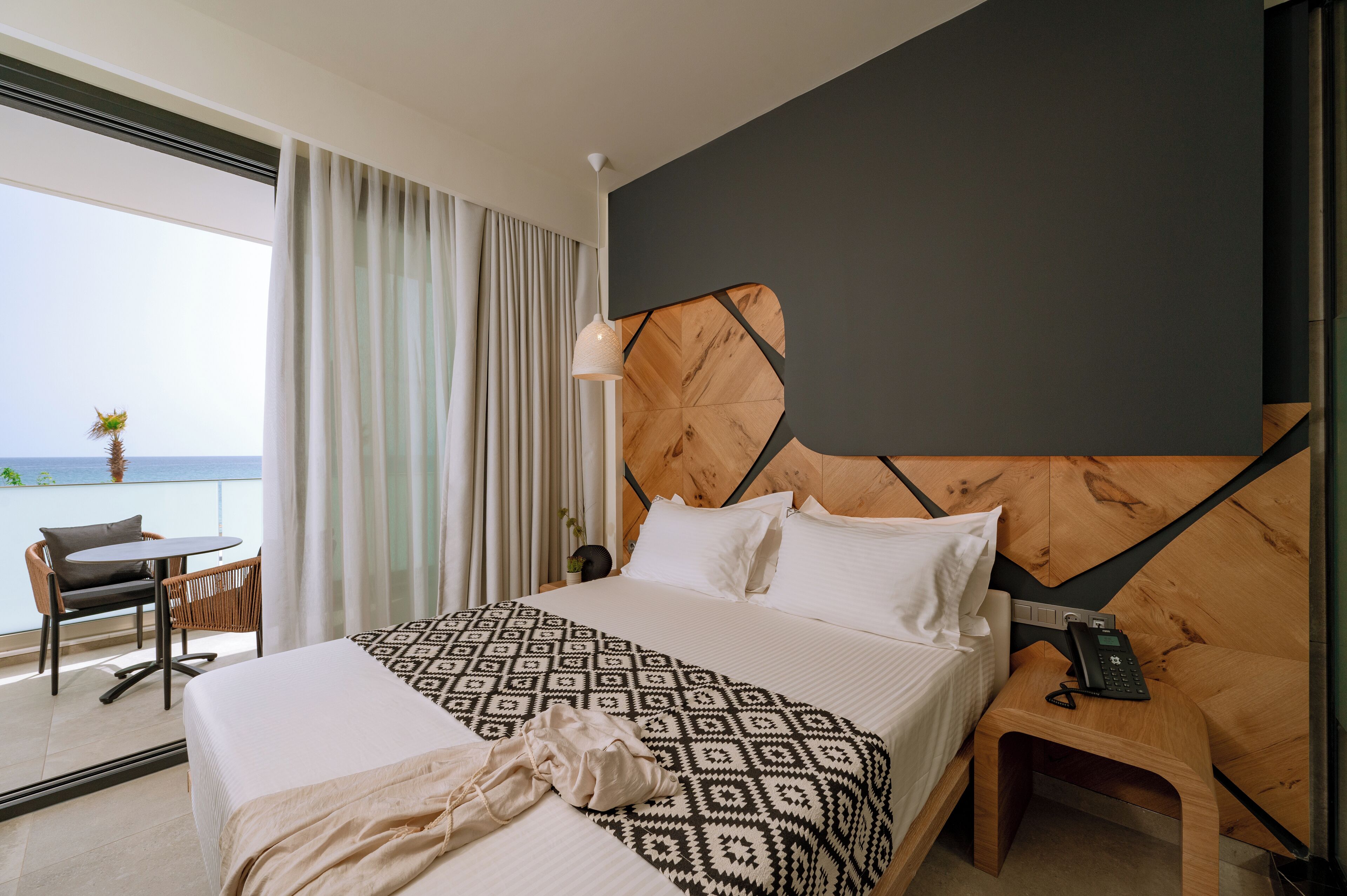Quarto de hotel com uma cama grande, cabeceira de madeira, mesa de cabeceira com telefone, pequena mesa com cadeiras e vista para o mar através de grandes janelas.