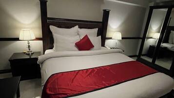 Cama bem arrumada com roupa de cama branca e vermelha, duas mesas de cabeceira com abajures e um espelho grande na parede.