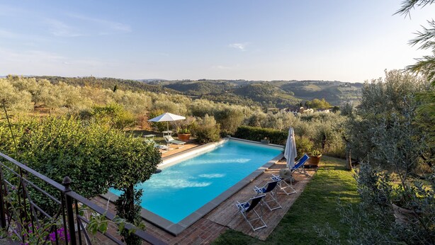 Uma piscina retangular com um deck de madeira e espreguiçadeiras, cercada por vegetação exuberante e colinas ao fundo.
