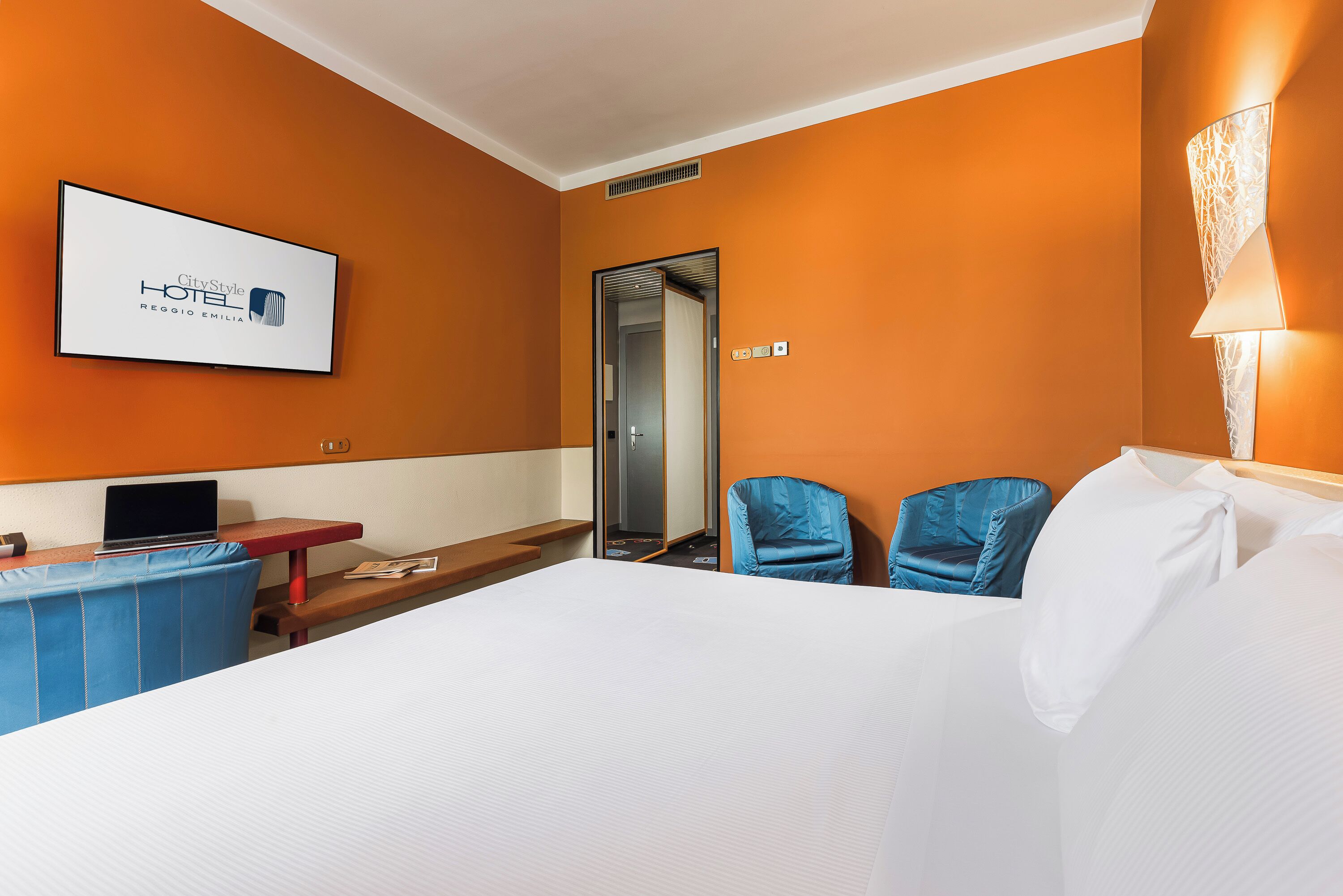 Quarto de hotel com uma cama, uma mesa com um laptop, duas cadeiras azuis e uma TV na parede.