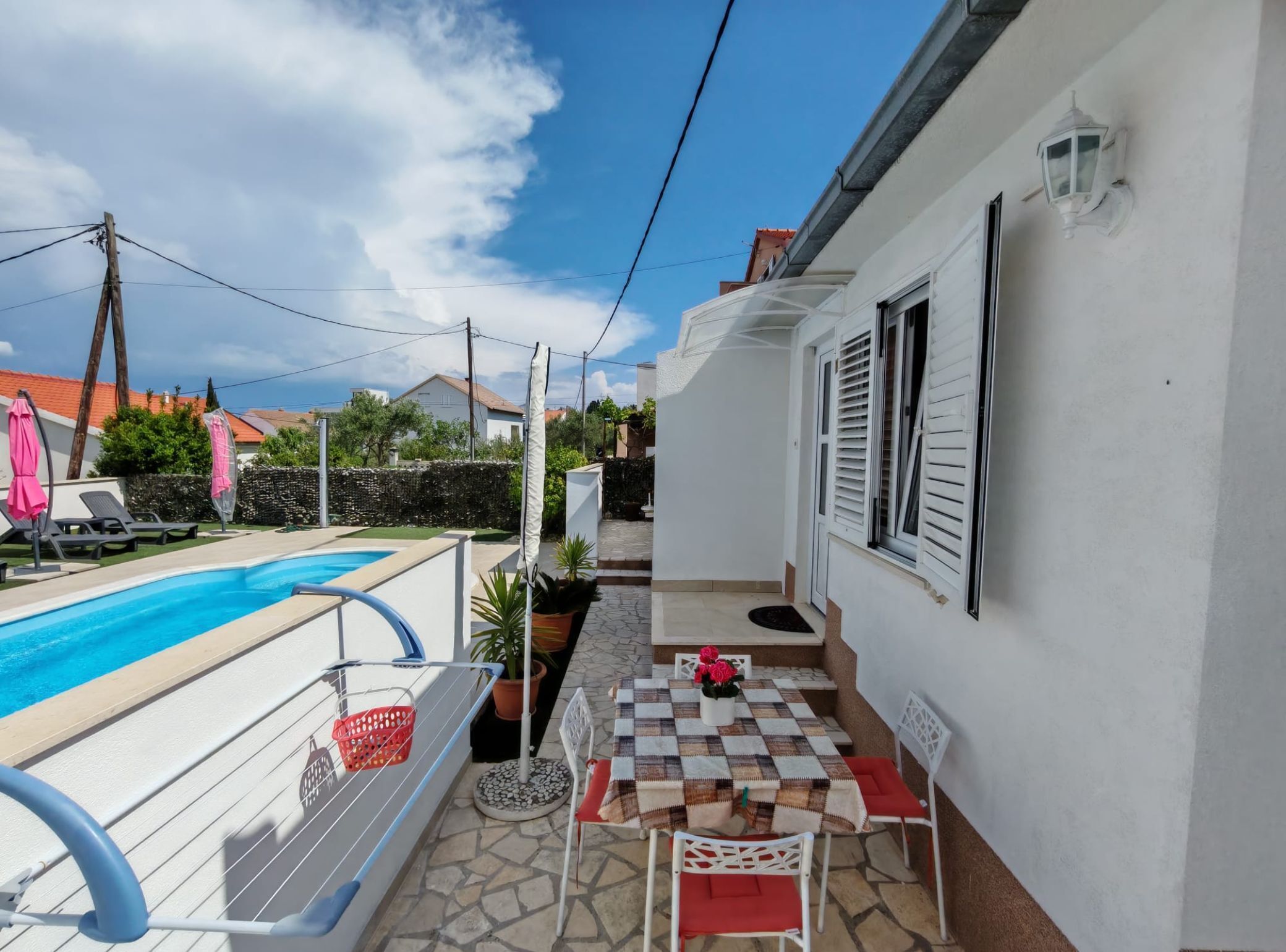 Uma área à beira da piscina com mesa e cadeiras, um pequeno jardim e um prédio branco com persianas.