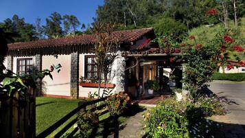 Uma pequena casa de pedra com telhado de cerâmica, rodeada por vegetação exuberante e plantas floridas.