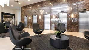 Um lobby moderno com cadeiras de couro preto, uma mesa redonda preta e uma parede de mármore com elevadores.