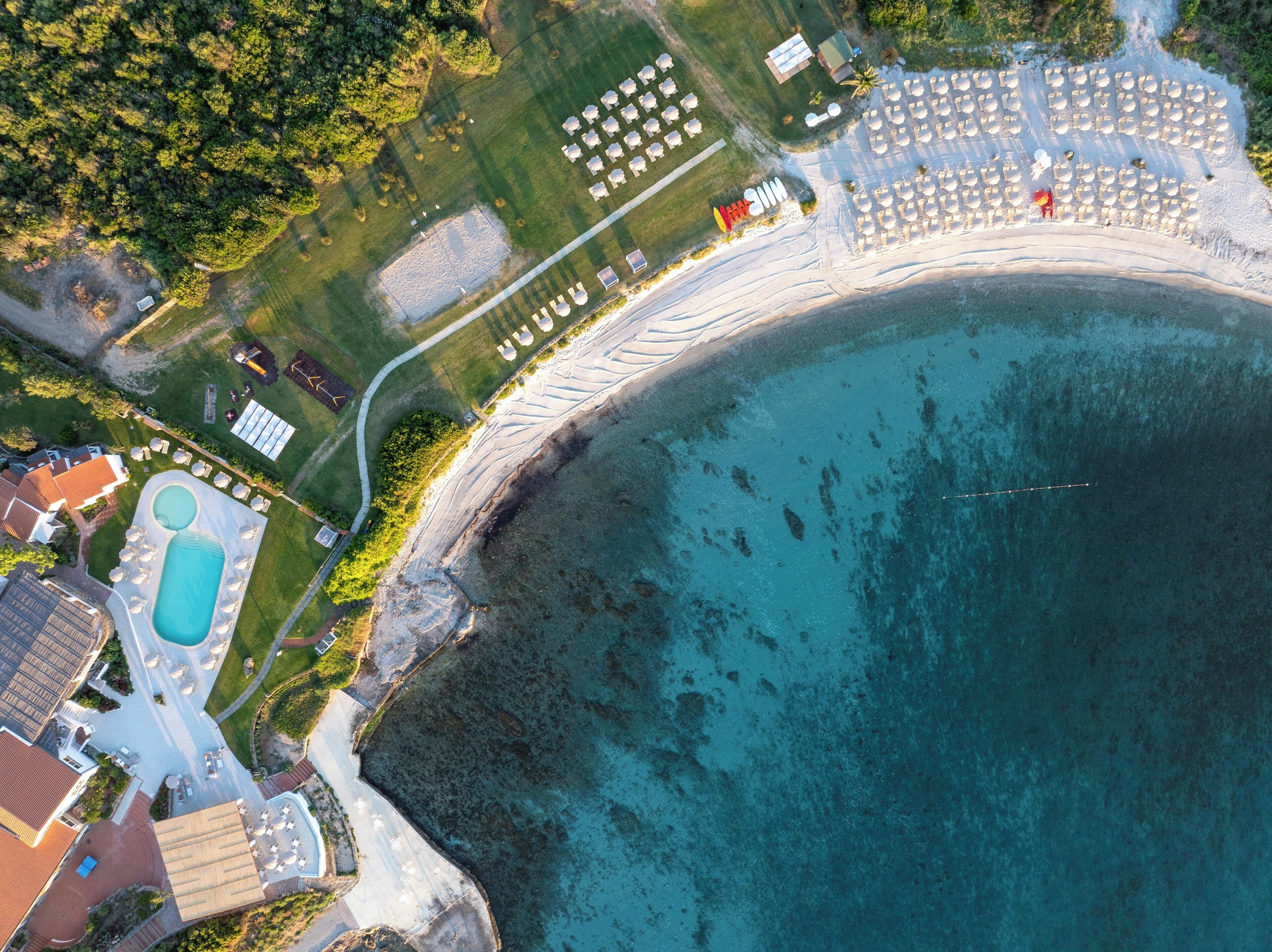 Vista aérea de uma propriedade costeira com piscina, guarda-sóis de praia e área de estacionamento.