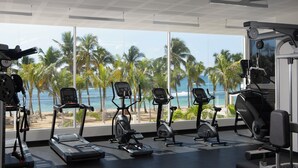 Academia com esteiras e bicicletas ergométricas, com vista para uma praia com palmeiras e o oceano.