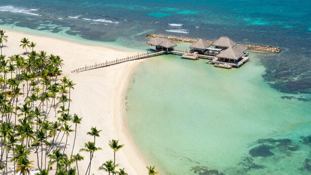 Vista aérea de um resort à beira-mar com bangalôs sobre a água, uma praia de areia e águas turquesa cristalinas.