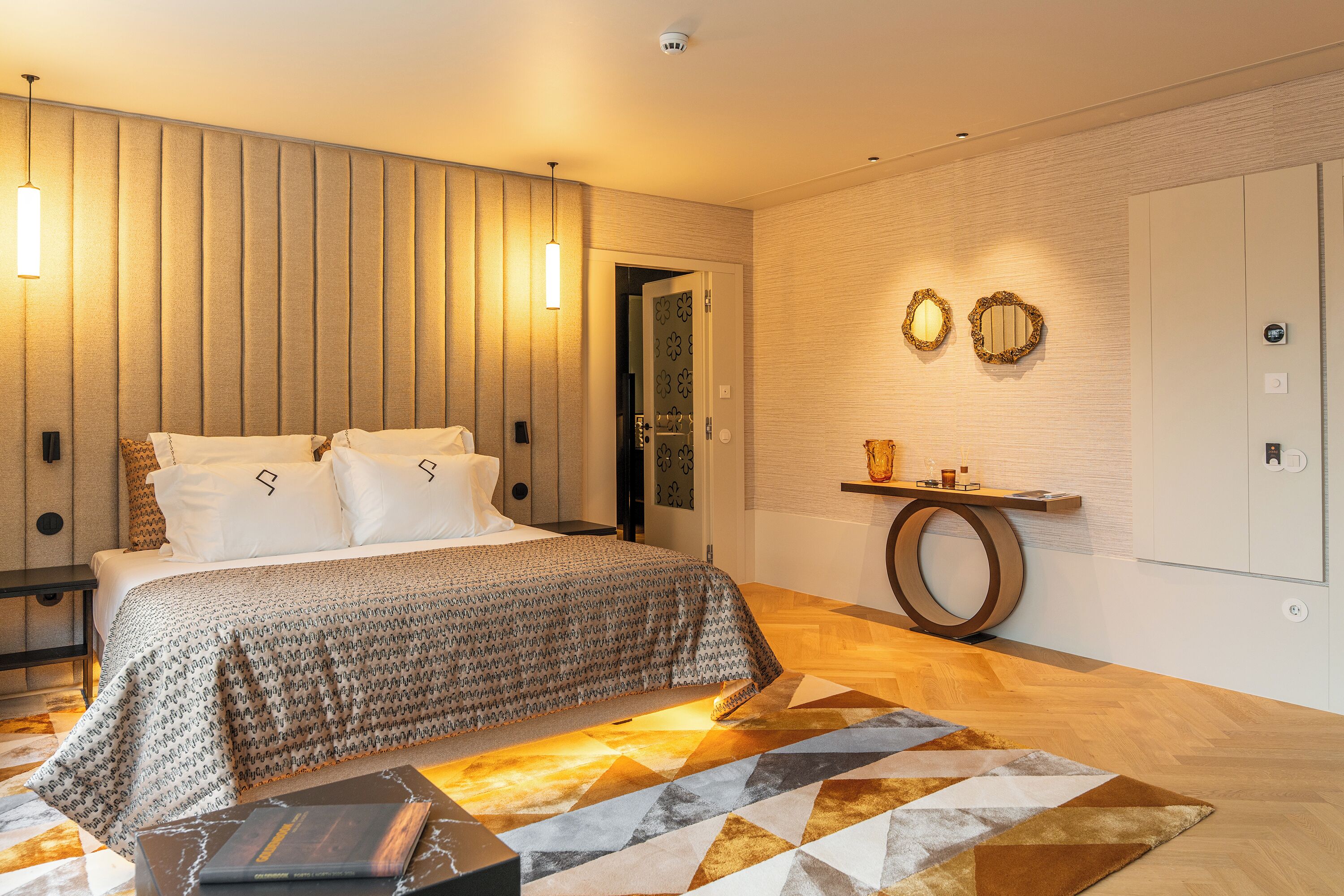 Quarto de hotel moderno com uma cama grande, uma mesa lateral redonda e um tapete com padrão geométrico.