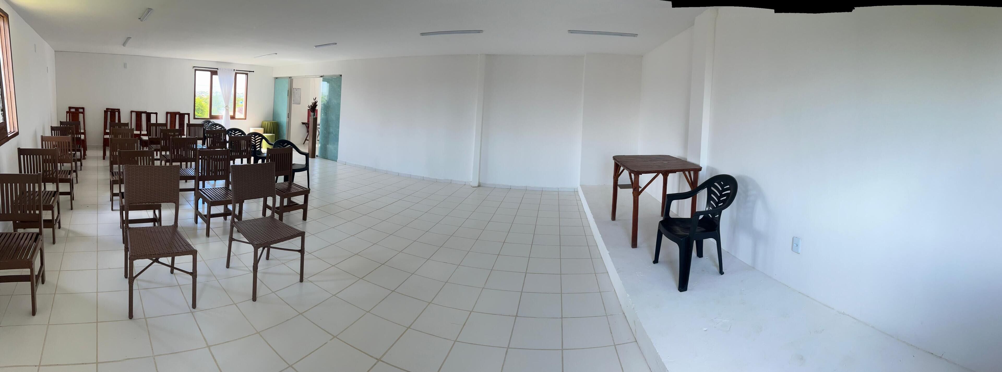Um quarto espaçoso com piso de azulejos, várias cadeiras e mesas de madeira, e uma grande janela que permite entrada de luz natural.