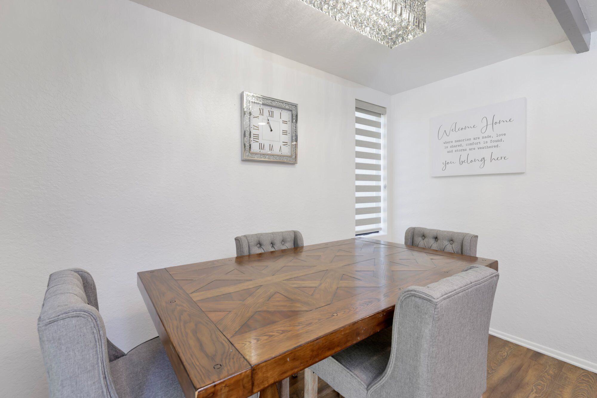 Uma sala de jantar com uma mesa de madeira, quatro cadeiras estofadas cinzentas, um relógio de parede e uma citação emoldurada.