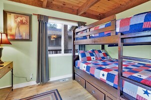 Uma cama beliche com colcha de retalhos, uma escrivaninha de madeira com luminária, uma janela com cortinas e um quadro na parede.