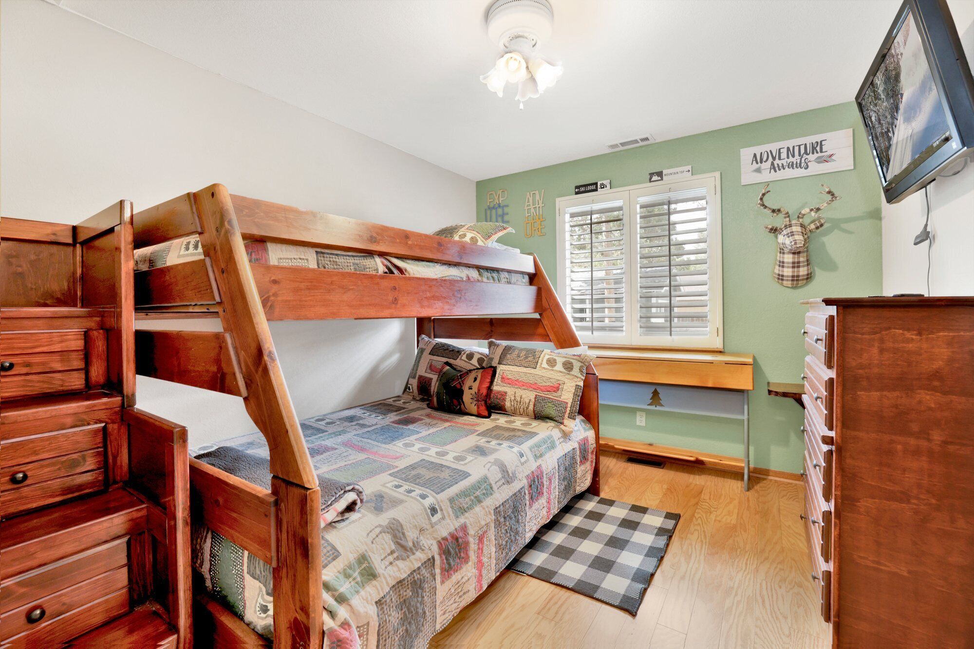Uma cama de beliche com colcha acolchoada, uma cômoda de madeira, uma janela com persianas e um painel com a inscrição 'Adventure Awaits'.