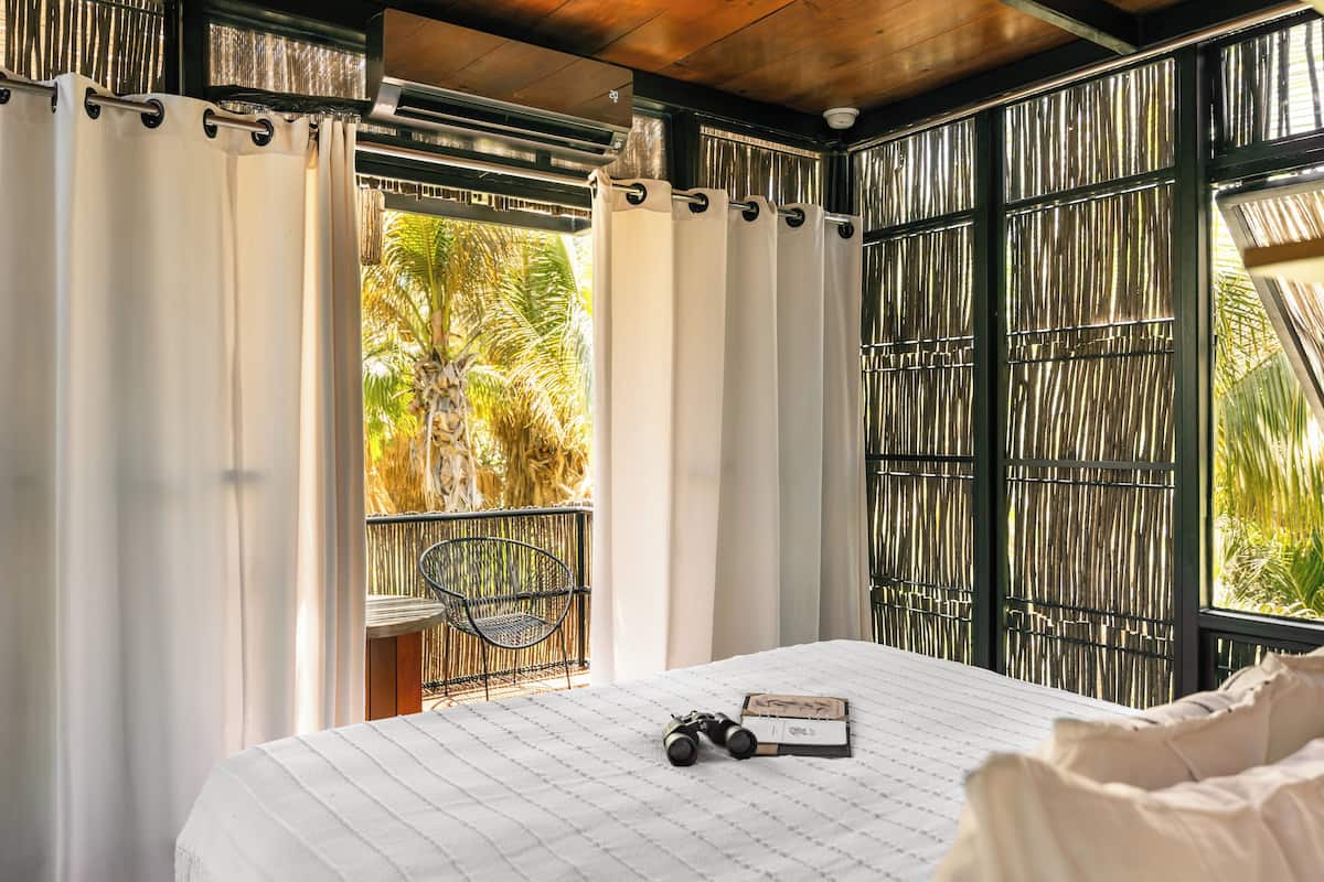 Um quarto com uma cama, um par de binóculos e um livro sobre a cama. Há uma janela com cortinas e vista para palmeiras lá fora.