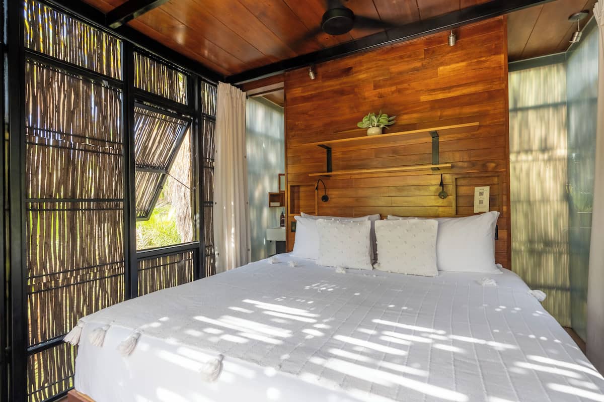 Uma cama grande com cabeceira de madeira, um vaso com planta e uma janela com cortinas de bambu.