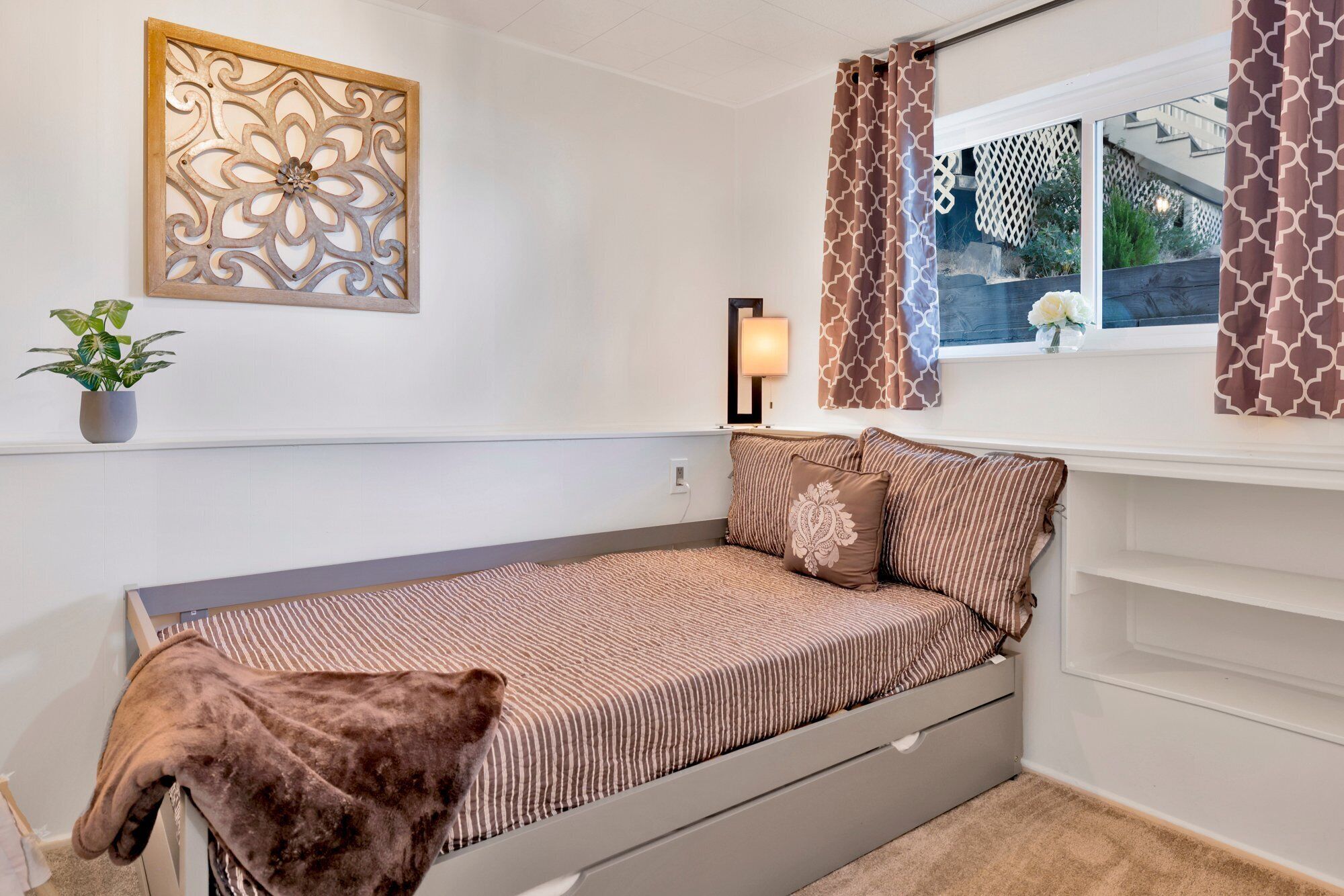 Quarto pequeno e bem arrumado, com cama, mesa de cabeceira, abajur, um objeto decorativo na parede e uma janela com cortinas.