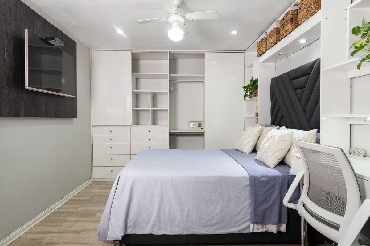 Quarto com cama, escrivaninha, cadeira, guarda-roupa e ventoinha no teto.