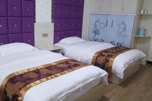 Quarto de hotel com duas camas, cabeceira roxa, guarda-roupa com obras de arte e um armário com chaleira.