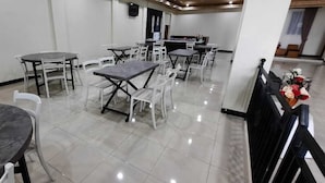 Uma área de refeições com várias mesas e cadeiras, uma cozinha ao fundo e uma escada que leva a um andar superior.