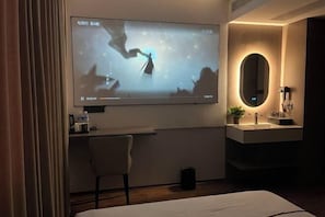 Quarto de hotel com uma televisão de tela grande fixada na parede, uma mesa de cabeceira com abajur, uma cadeira e uma cama.