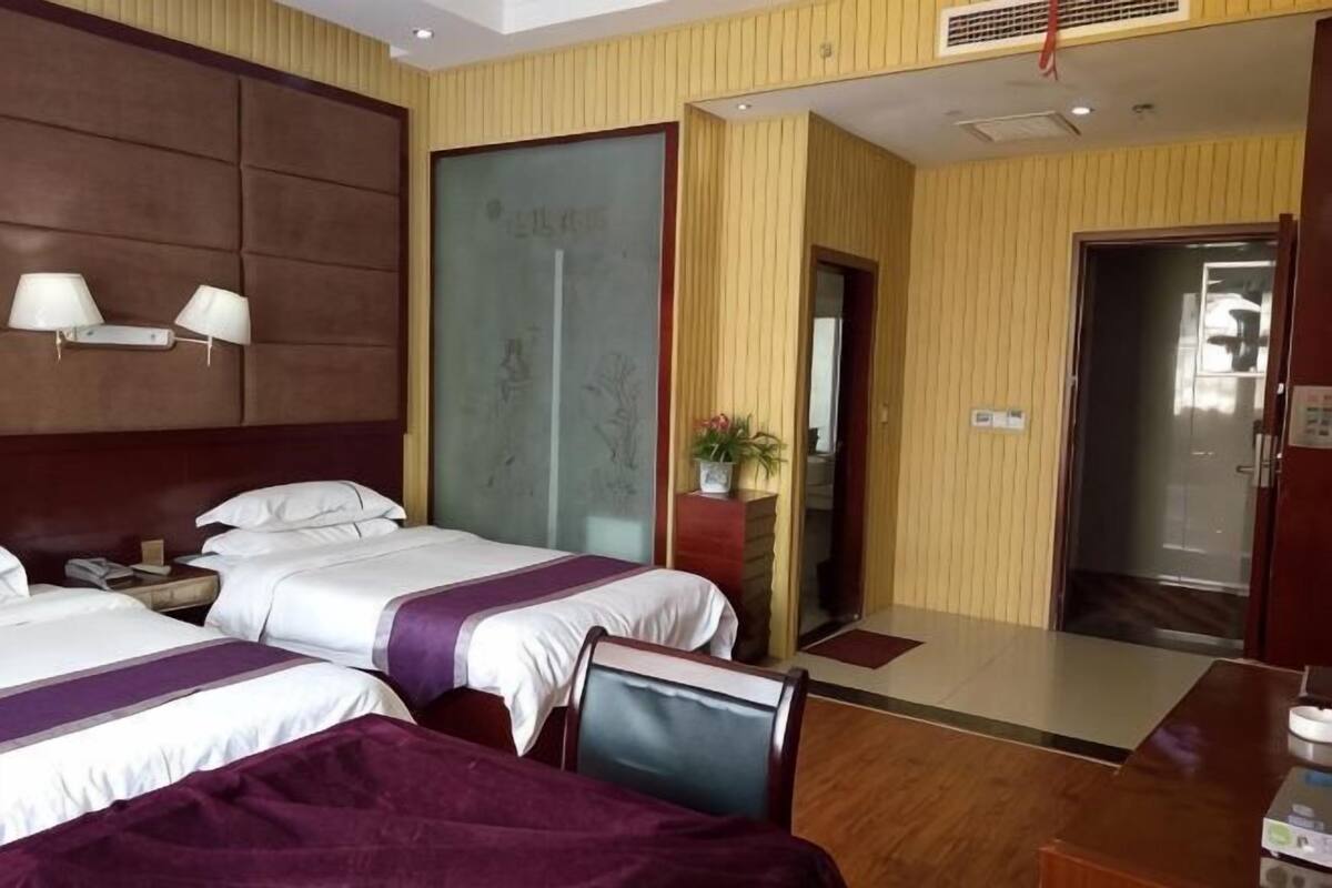 Quarto de hotel com duas camas, uma escrivaninha, uma cadeira, uma planta e uma porta.