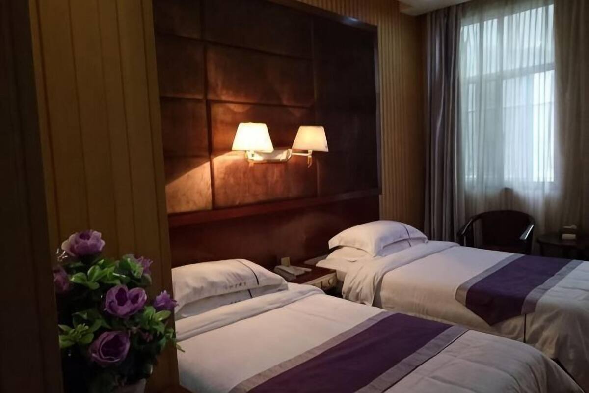 Quarto de hotel com duas camas, cabeceira de madeira, luminárias de cabeceira, uma cadeira e uma janela com cortinas.