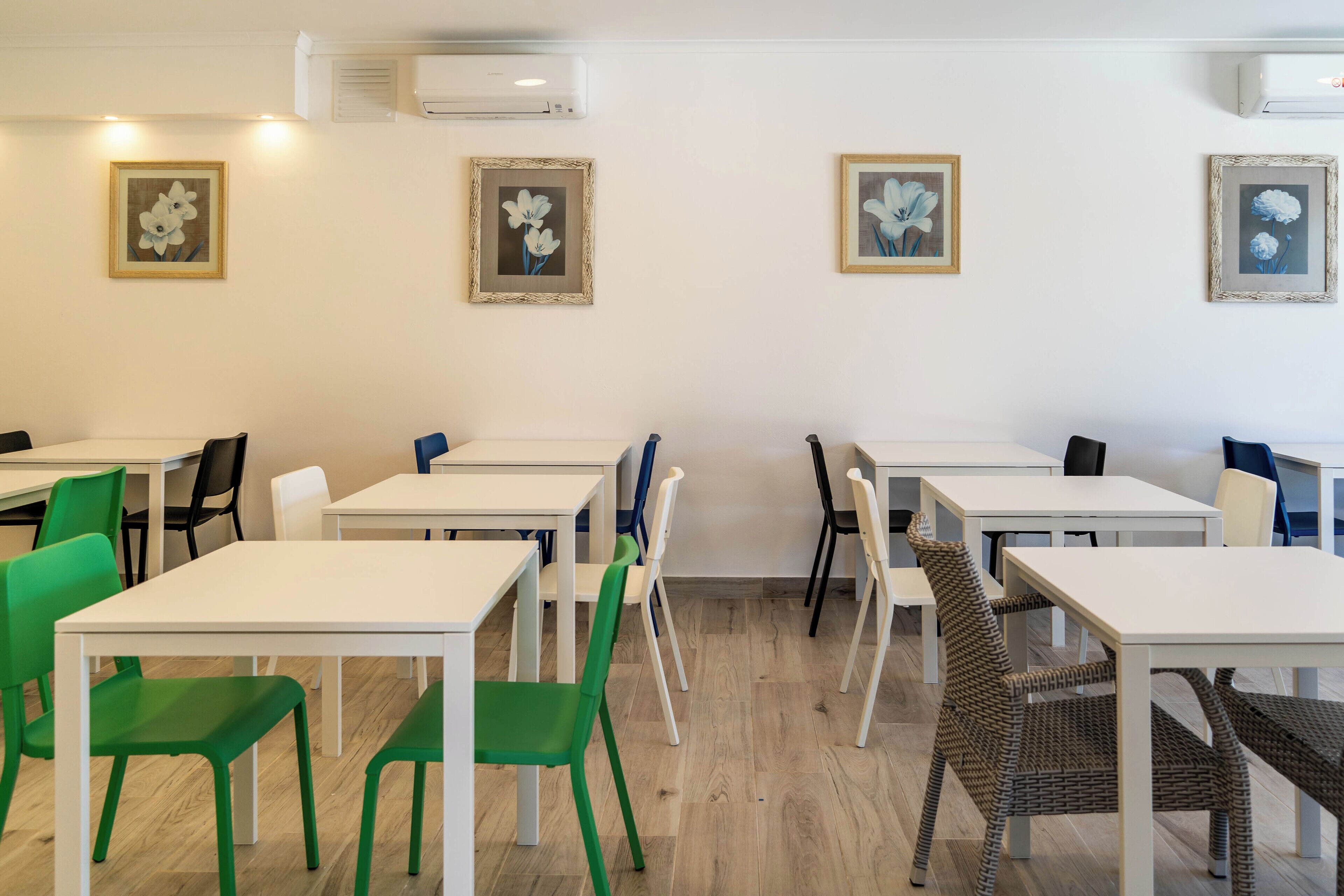 Um espaço com mesas e cadeiras brancas, algumas verdes e de vime, dispostas em configuração de sala de aula ou sala de reuniões.