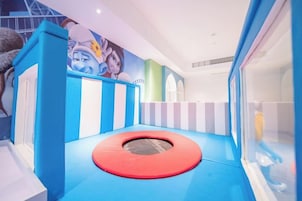 Espaço de recreação infantil com trampolim, paredes acolchoadas coloridas e um mural na parede de fundo.